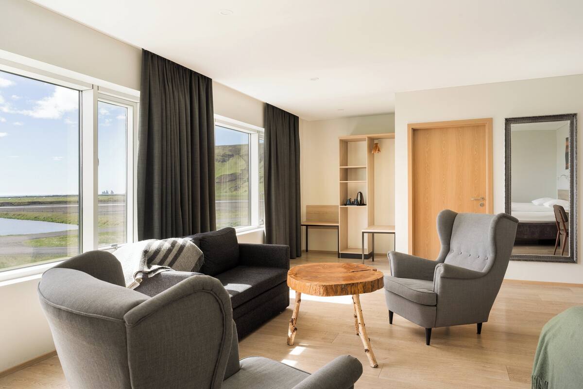 Sala de estar moderna com sofá, poltrona e mesa de madeira redonda. Uma grande janela oferece vista para a paisagem.
