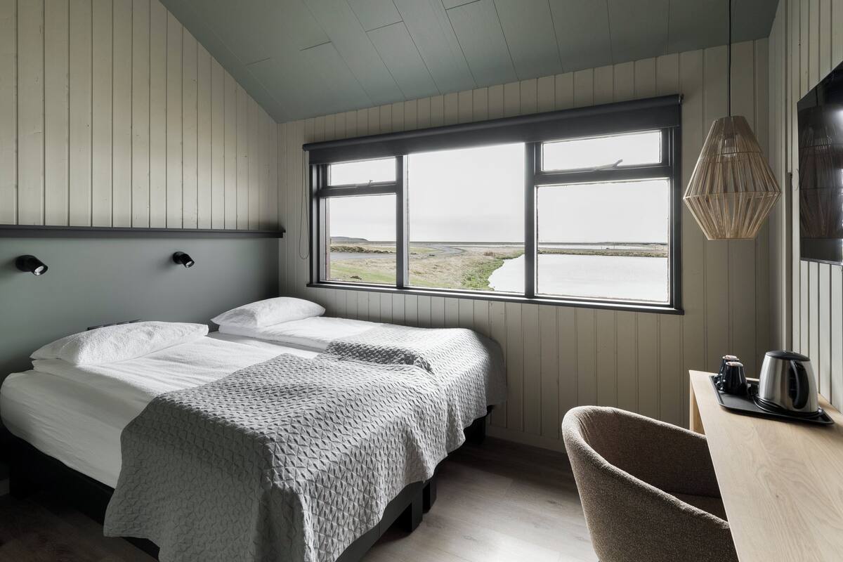 Quarto com cama, poltrona, escrivaninha com chaleira e vista para o mar atrav és de grandes janelas.