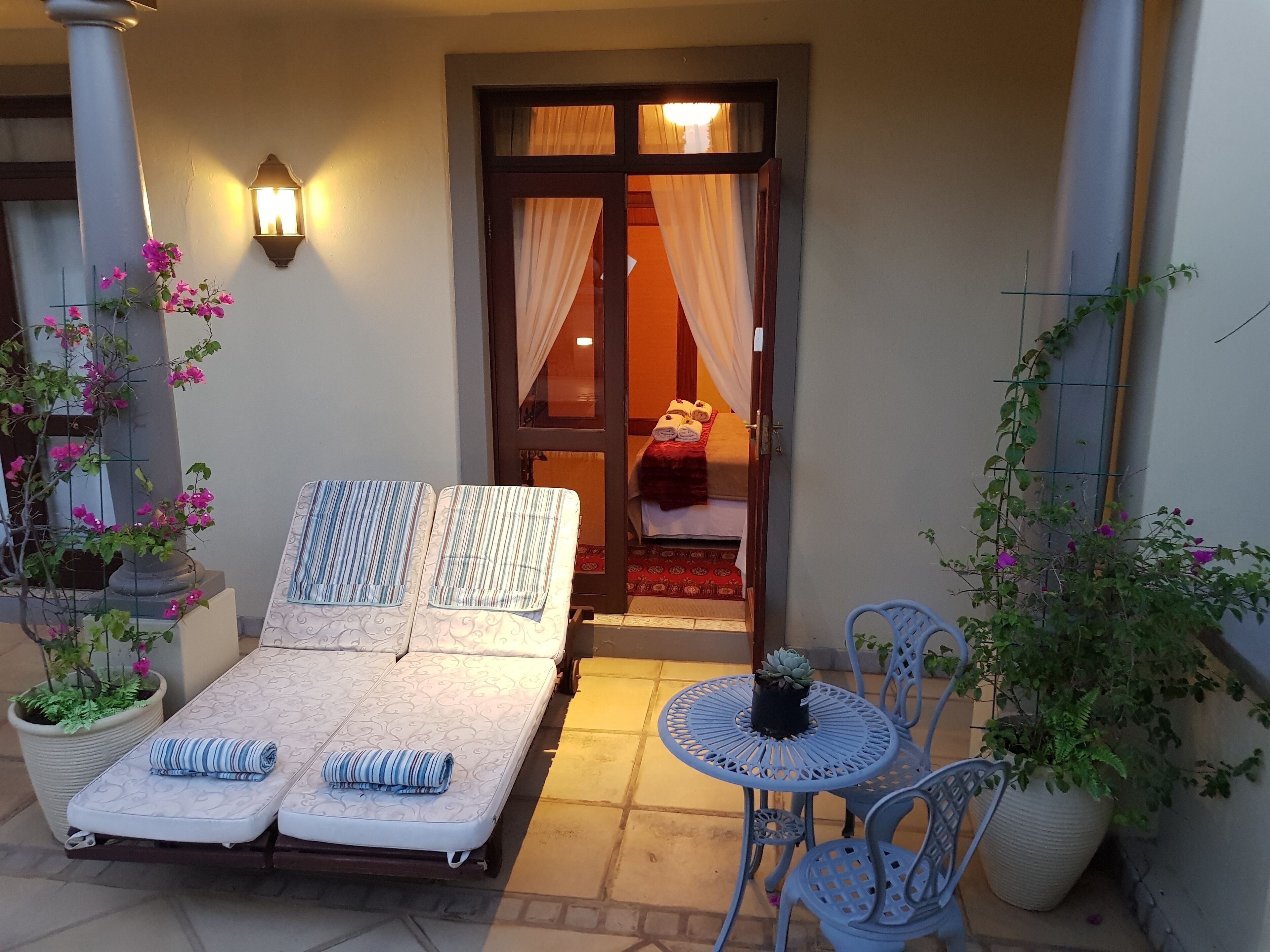 Área externa ensolarada com assentos, incluindo um sofá e cadeiras, uma mesa pequena com uma planta, e uma porta que dá para um quarto com cama e toalhas.