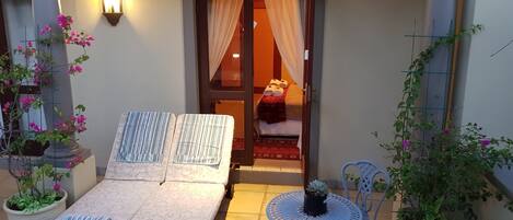 Área externa ensolarada com assentos, incluindo um sofá e cadeiras, uma mesa pequena com uma planta, e uma porta que dá para um quarto com cama e toalhas.