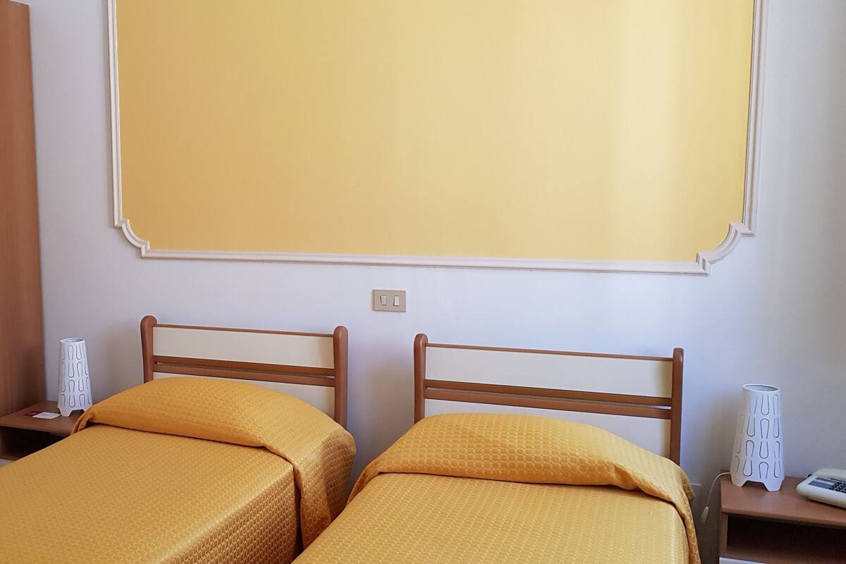 Quarto com duas camas, cabeceiras de madeira, uma mesinha de cabeceira com abajur e uma janela com persiana amarela.