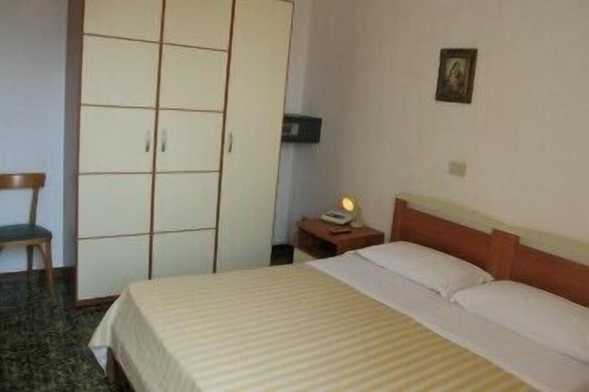 Quarto de hotel com cama, guarda-roupa, cadeira, mesa de cabeceira com luminária e um quadro na parede.