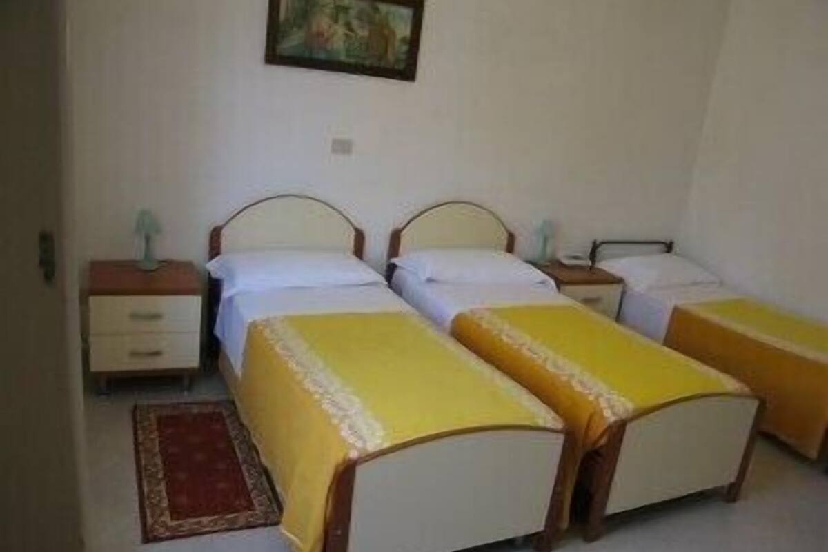 Quarto com duas camas de solteiro, cada uma com cabeceira de madeira e colcha amarela. Em cada lado das camas, há uma mesinha de cabeceira com uma luminária, e um pequeno canto com uma mesa e uma cadeira.