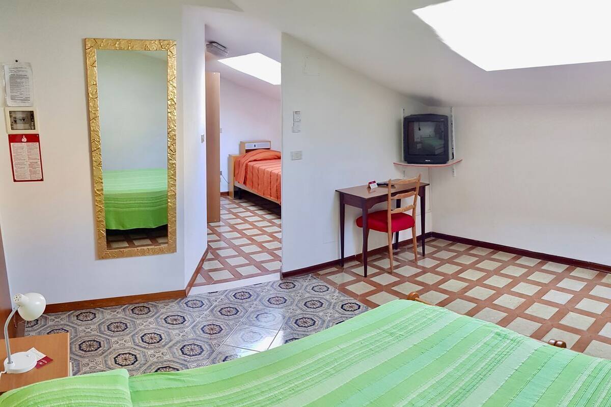 Quarto de hotel com cama, escrivaninha com cadeira, televisão e piso estampado.