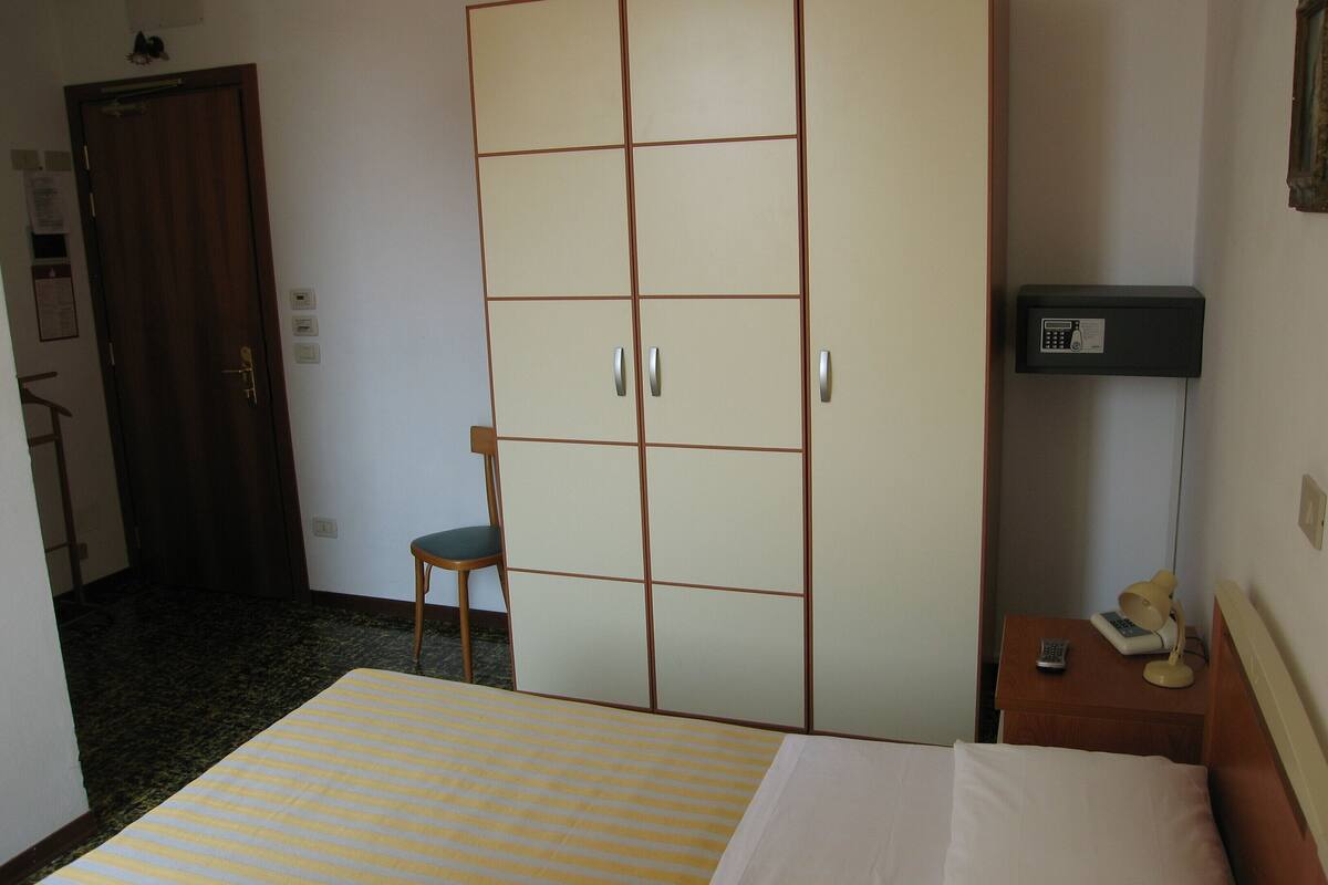 Quarto de hotel com cama, guarda-roupa, cadeira, mesa de cabeceira com abajur e telefone.