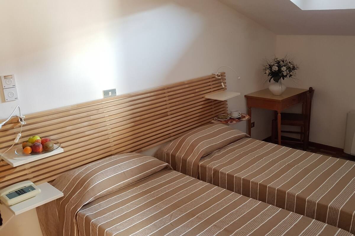 Quarto de hotel com duas camas, uma mesa de cabeceira com abajur, uma mesinha com um vaso de flores e um prato de frutas na cabeceira.