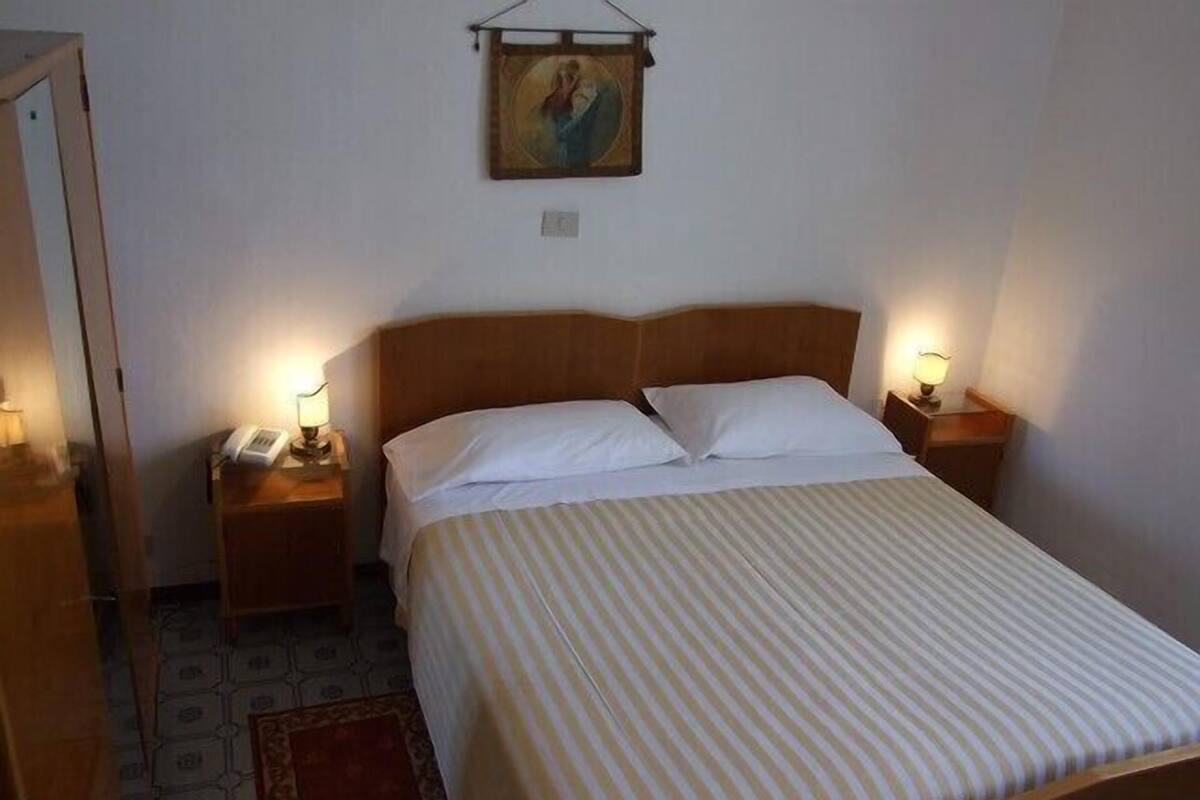 Quarto de hotel com uma cama de solteiro, duas mesas de cabeceira com luminárias, uma cabeceira de madeira, um tapete estampado e um quadro na parede.