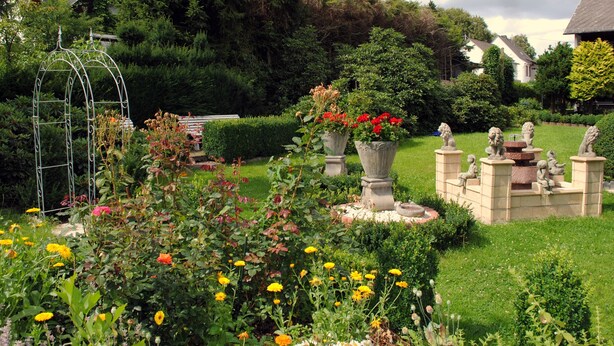 Um jardim com treliça de metal, canteiros de flores e esculturas de pedra.