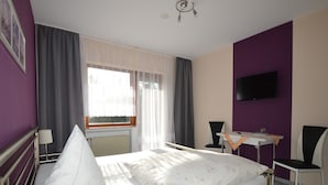 Quarto de hotel com cama, secretária, cadeira, televisão e janela com cortinas.