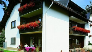 Um edifício de dois andares com varandas decoradas com flores vermelhas, uma área de estar coberta ao ar livre e um céu claro.