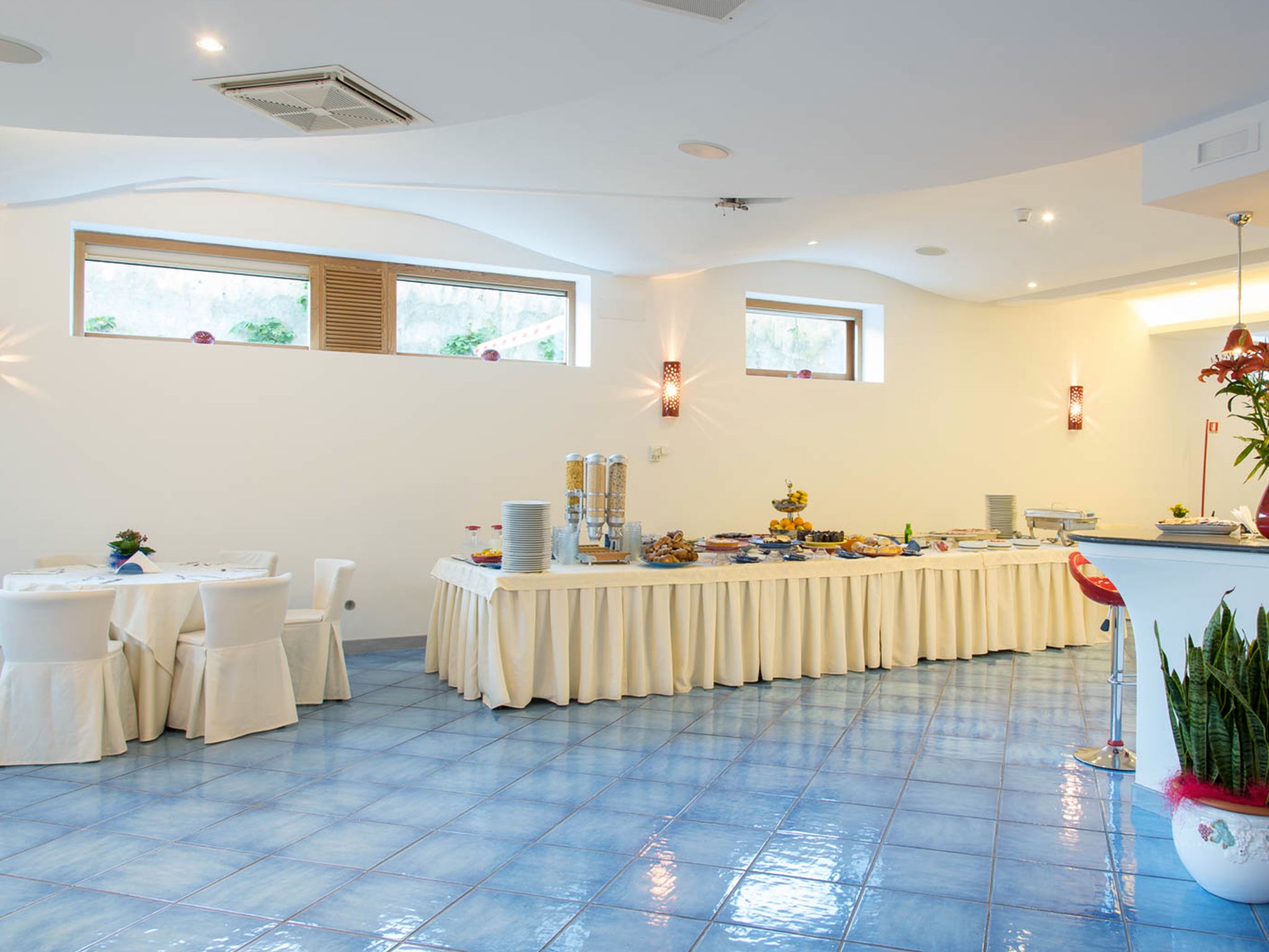Um quarto amplo com uma mesa comprida preparada para um buffet, cadeiras brancas e grandes janelas com persianas.