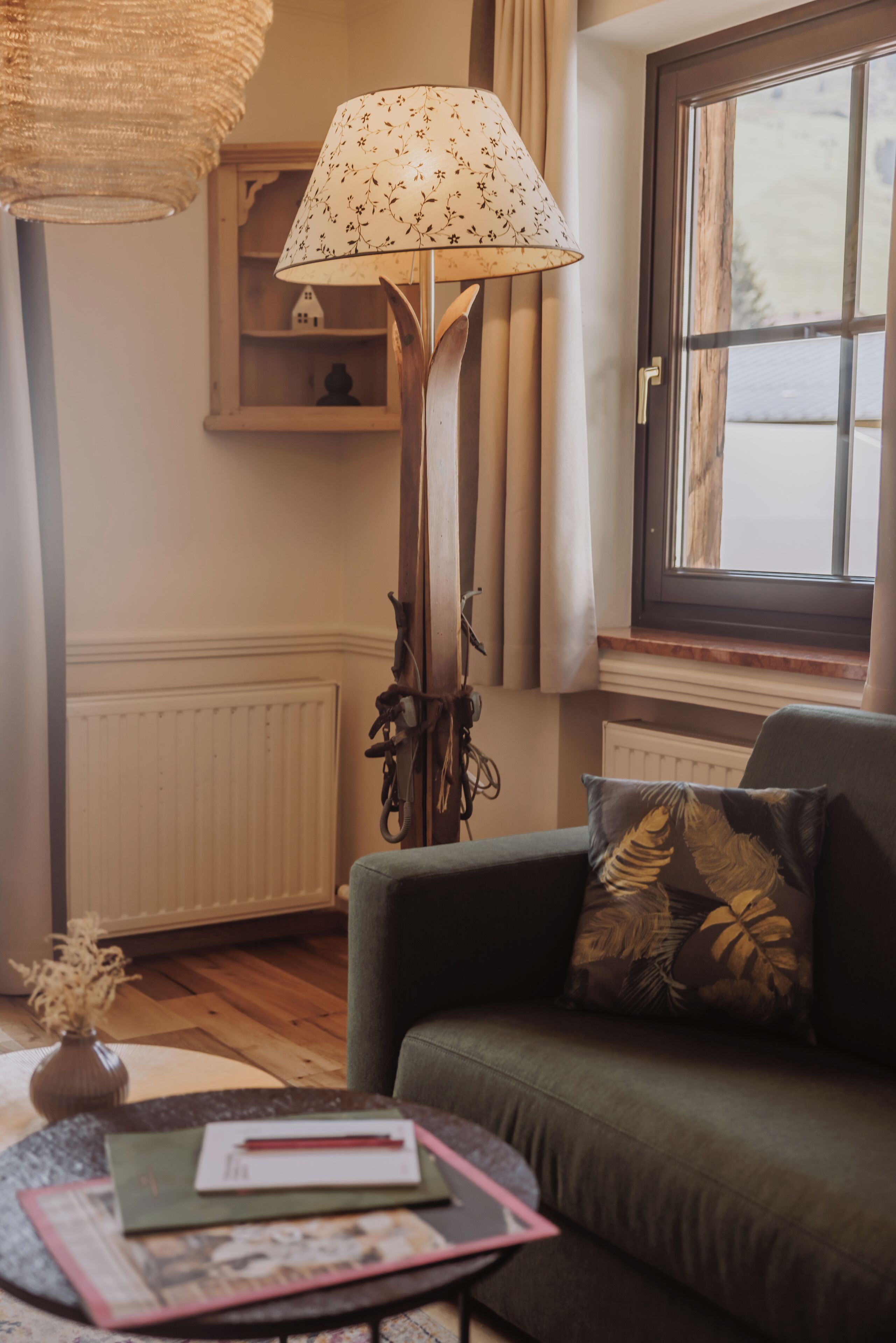 Uma sala de estar aconchegante com sofá, luminária de chão, mesinha com livros, um vaso com flores secas e uma janela com cortinas.