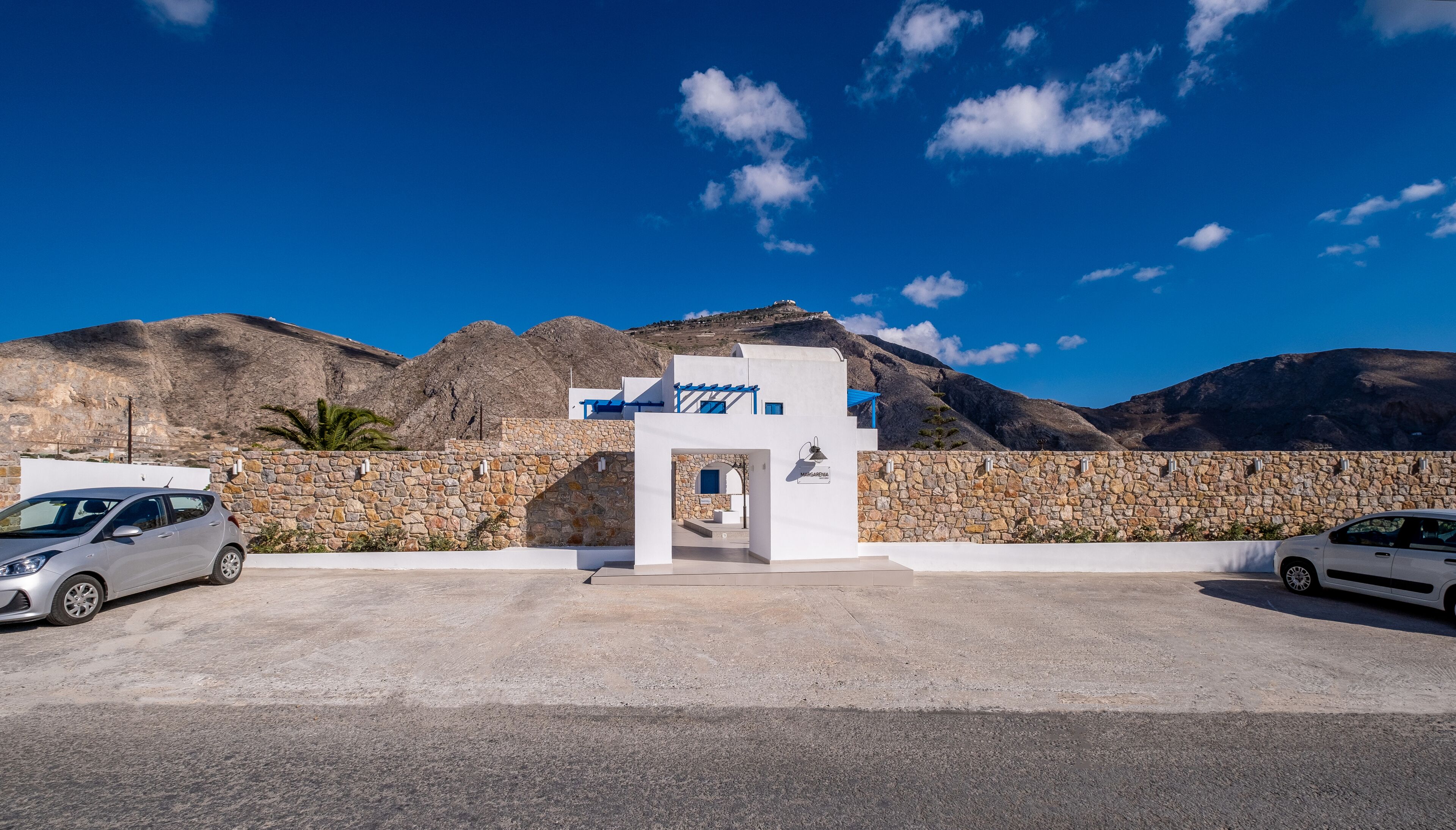 Um edifício branco com entrada de parede de pedra, céu azul límpido e montanhas ao fundo.