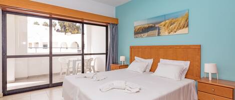 Quarto de hotel com uma cama grande, cabeceira de madeira, um quadro na parede e vista através da porta de vidro.