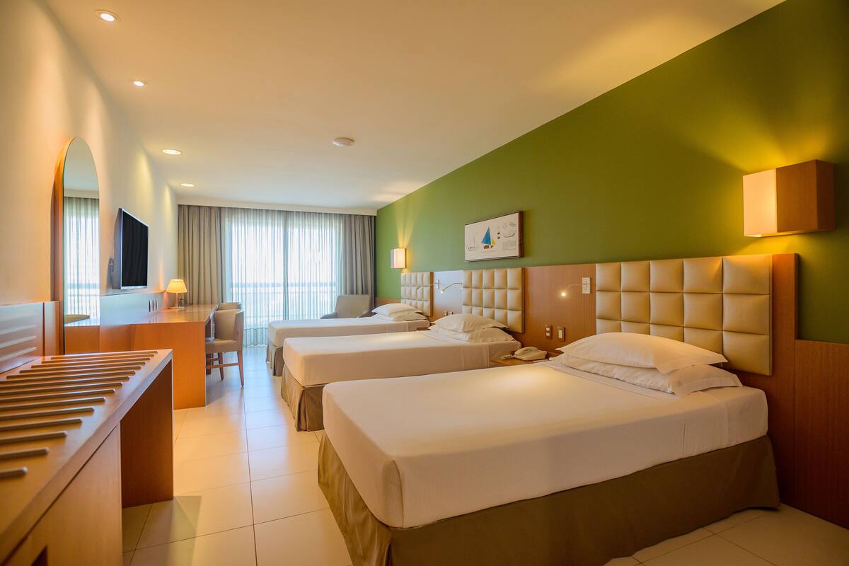 Quarto de hotel com três camas, uma escrivaninha, televisão e uma janela grande com cortinas.