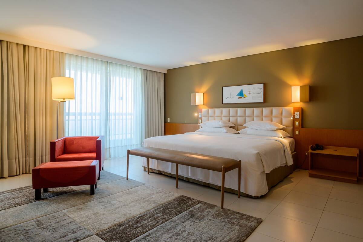 Quarto de hotel com uma cama grande, uma poltrona vermelha, um banco e uma mesinha de cabeceira.