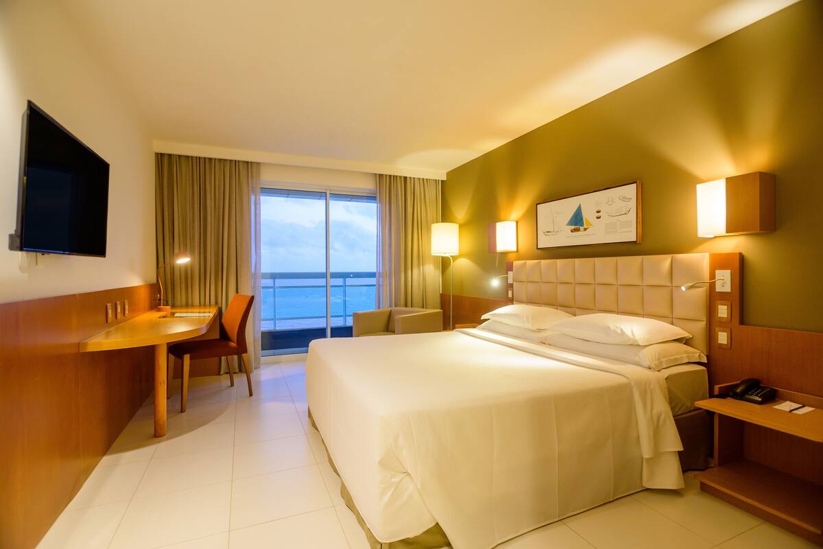 Quarto de hotel com cama, escrivaninha, cadeira, televisão e vista para o mar.