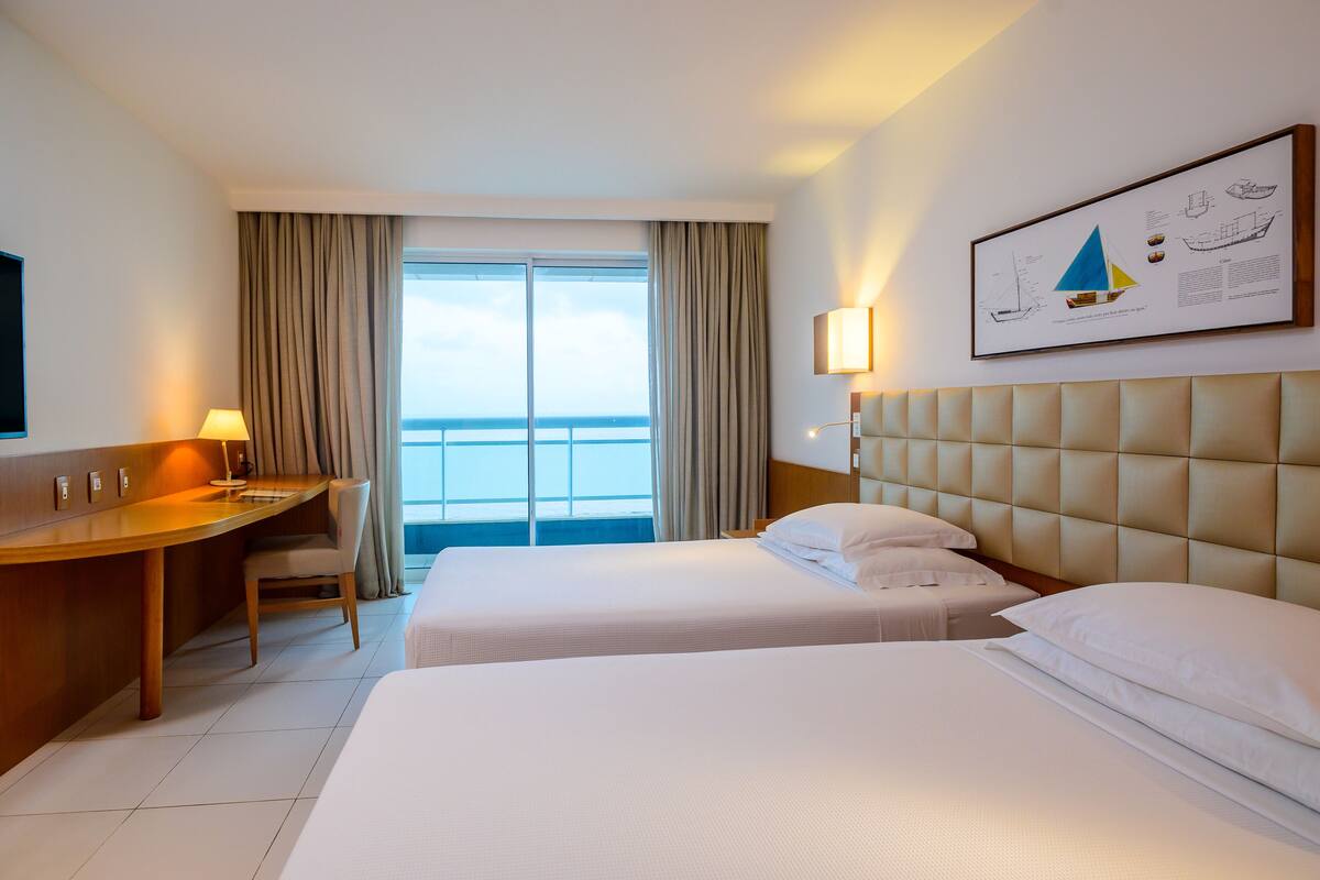 Quarto de hotel com duas camas, uma escrivaninha, uma cadeira e uma janela grande com cortinas.