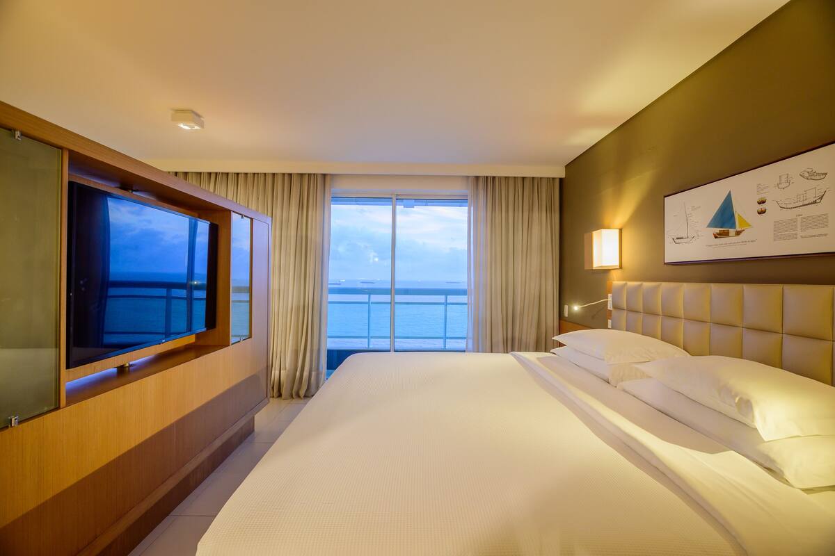 Quarto de hotel com uma cama grande, televisão, vista para o mar e um quadro pendurado na parede.