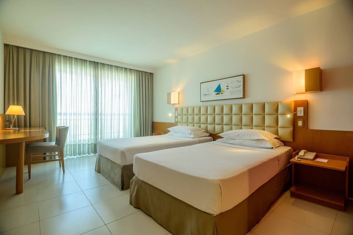 Quarto de hotel com duas camas, uma escrivaninha, uma cadeira e uma mesa de cabeceira com telefone.