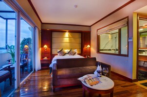 Quarto de hotel com uma cama, uma mesinha com uma revista e copos de vinho, um sofá e um espelho grande.