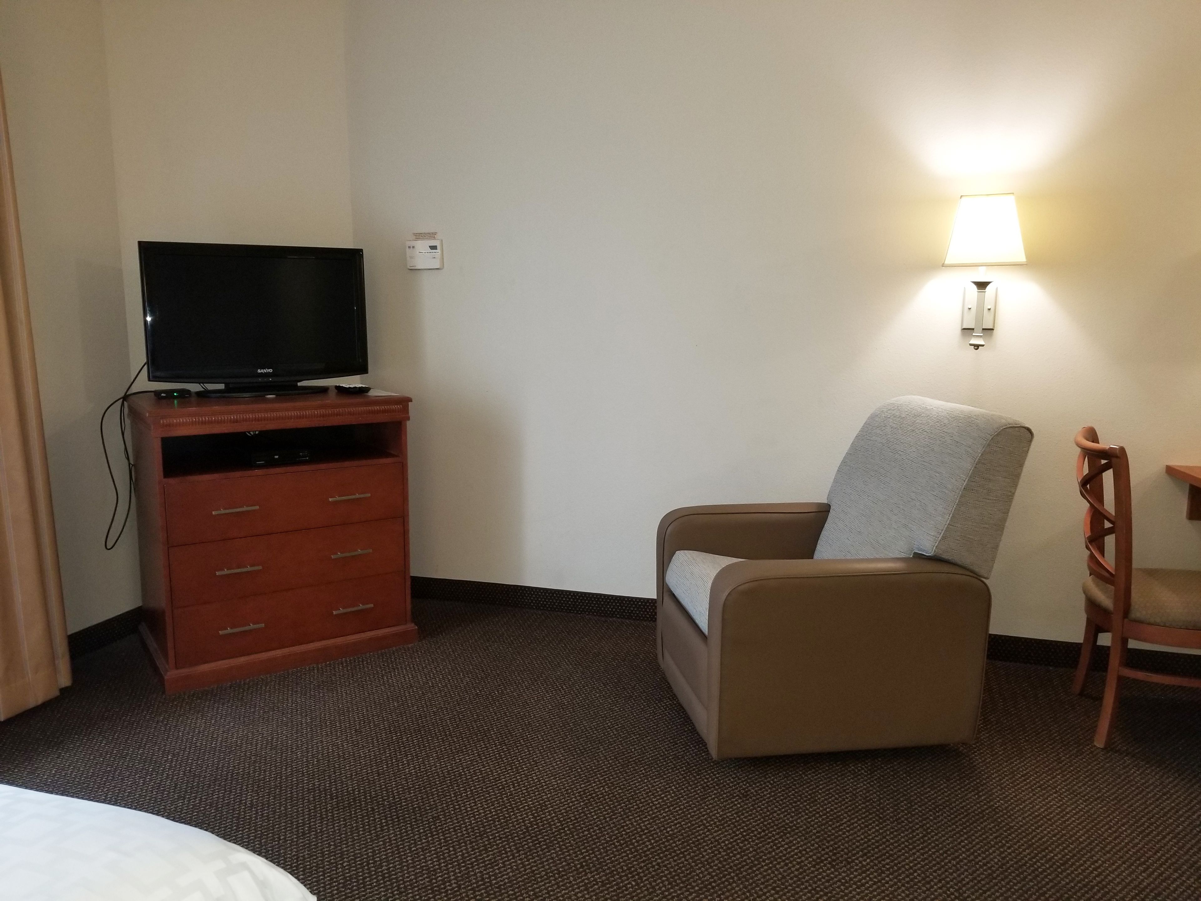 Quarto de hotel com televisão em móvel de madeira, uma cadeira, abajur e piso coberto por carpete.