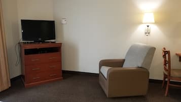 Quarto de hotel com televisão em móvel de madeira, uma cadeira, abajur e piso coberto por carpete.