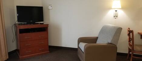 Quarto de hotel com televisão em móvel de madeira, uma cadeira, abajur e piso coberto por carpete.