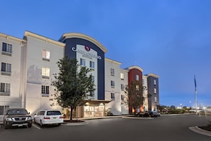 Um hotel com design moderno, exibindo uma placa bem visível da Candlewood Suites. Vários espaços de estacionamento são visíveis em frente ao edifício.