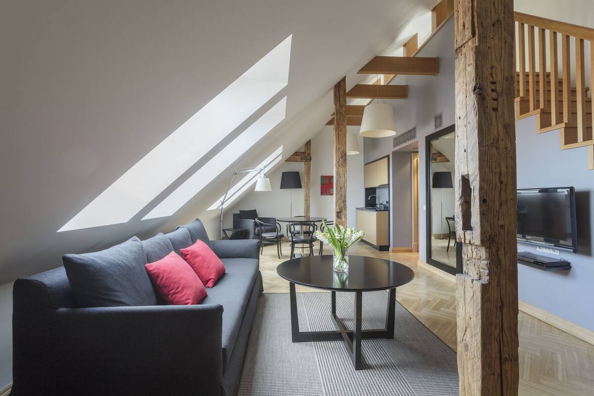 Um loft moderno com pé direito alto, claraboias, um sofá preto com almofadas vermelhas, uma mesa de centro redonda e uma televisão grande.