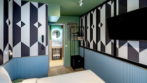 Quarto de hotel com cama, televisão fixada na parede, banheiro com espelho e prateleira com produtos de higiene pessoal.