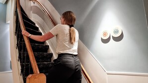 Uma pessoa descendo uma escadaria de madeira com parede estampada e duas placas circulares com números de quartos e um aviso de boas-vindas.