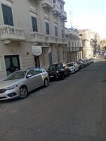 Fila de carros estacionados na rua, com edifícios que têm sacadas e placas.