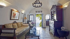 Uma sala de estar acolhedora com um sofá, uma mesa de centro e um candelabro. Há uma porta que dá para uma varanda com plantas.
