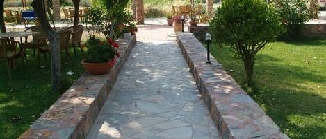 Um caminho de pedra leva a um espaço de estar ao ar livre sob uma pérgola, cercado por vasos com plantas e vegetação.