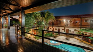 Um deck de madeira com piscina, cercado por plantas tropicais e um edifício com telhado em azulejos.