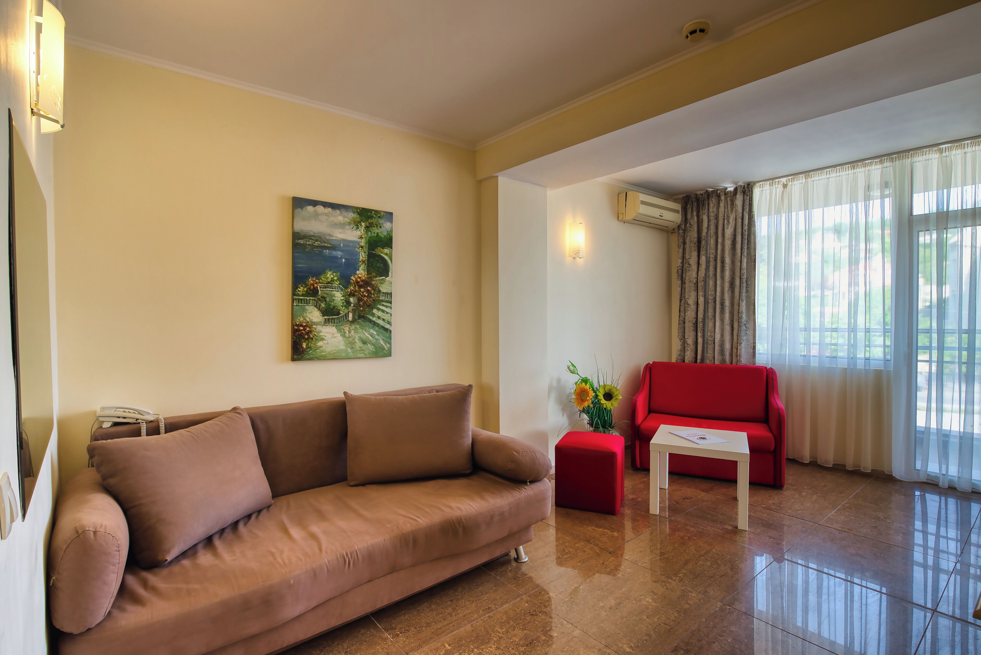 Uma sala de estar com sofá, uma poltrona vermelha, mesa de centro, quadro na parede e um ar condicionado.