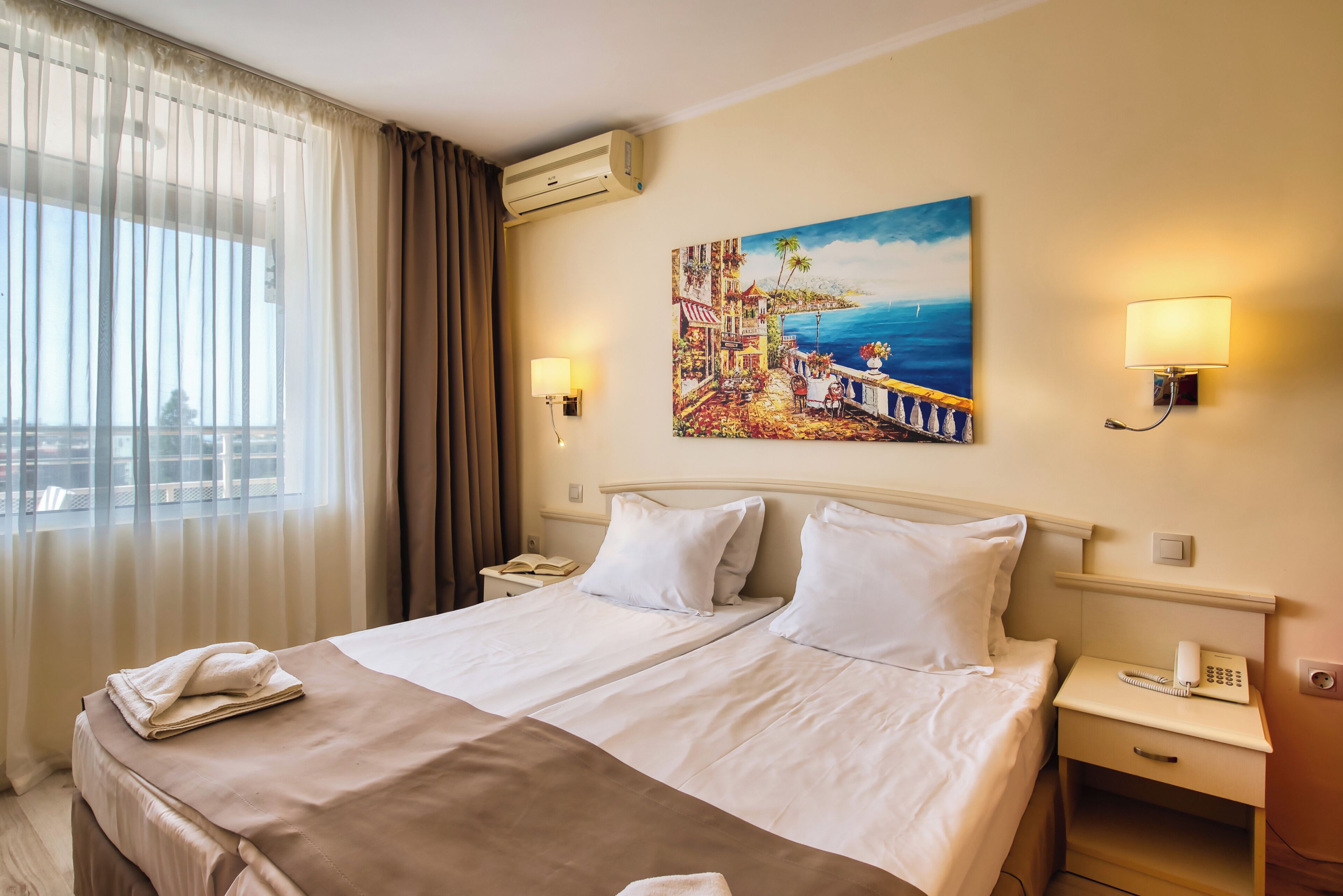 Quarto de hotel com cama, duas mesas de cabeceira, um criado-mudo, um quadro na parede e uma janela com cortinas.