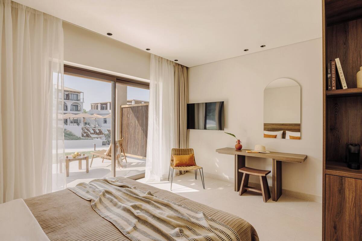 Quarto de hotel moderno com uma cama grande, uma escrivaninha e uma cadeira. Há uma varanda com vista para o hotel e arredores.