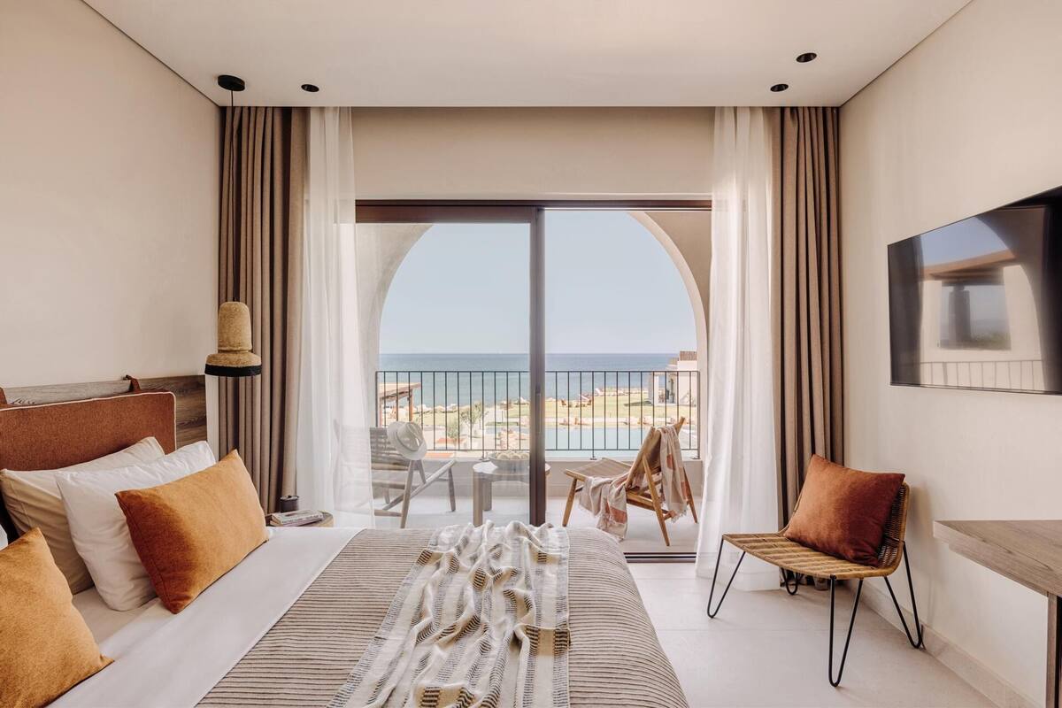 Quarto de hotel com uma cama grande, uma cadeira, uma mesa pequena e vista para o mar.
