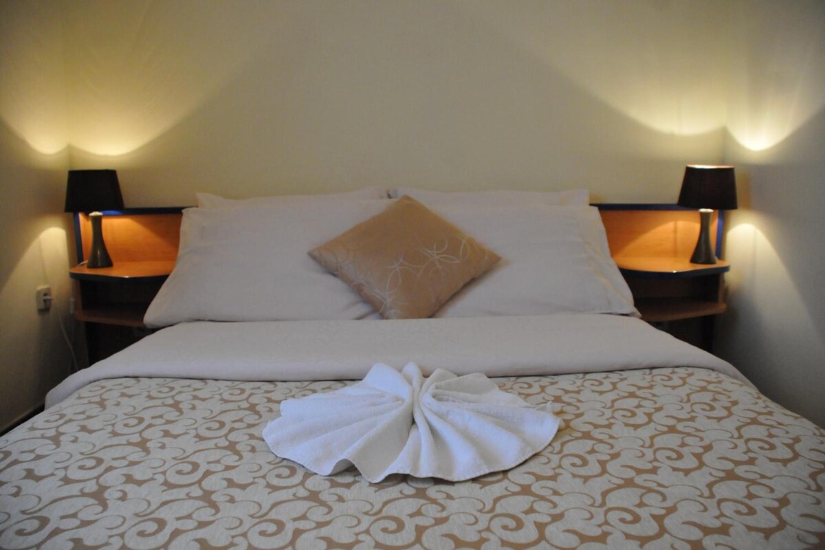 Quarto de hotel com cama bem arrumada, duas luminárias de cabeceira e um travesseiro decorativo.