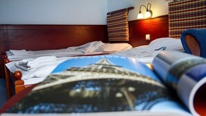 Quarto de hotel com cama bem arrumada, cabeceira de madeira, luminária na parede e toalha xadrez pendurada.