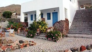 Um edifício branco com persianas azuis, uma escadaria de pedra e um jardim com diversas plantas e flores.