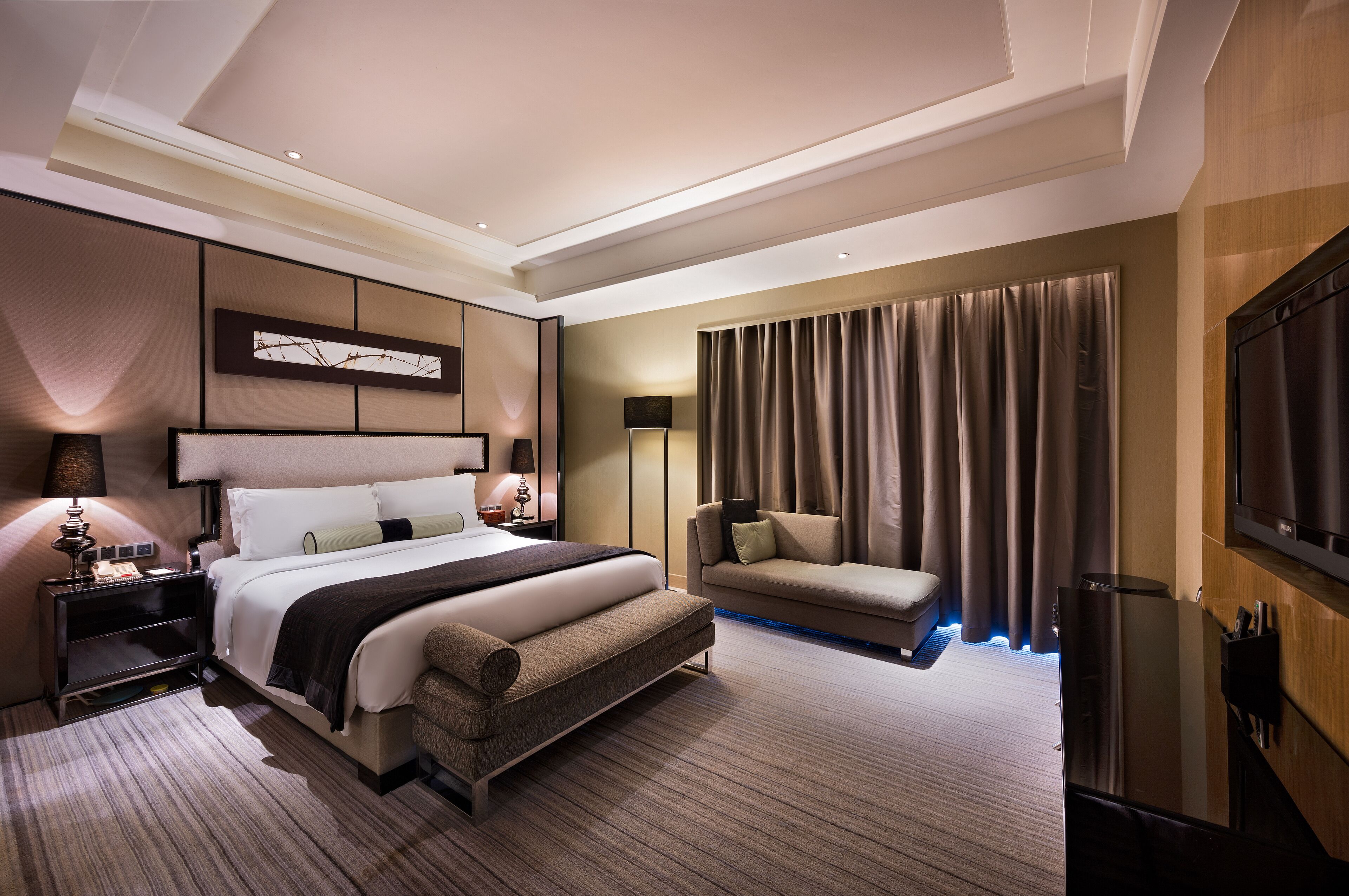 Quarto de hotel moderno com uma cama grande, um sofá, uma televisão de tela plana e uma janela ampla com cortinas.