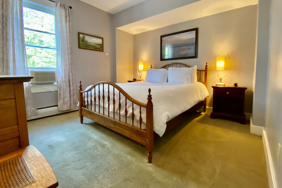 Um quarto com uma cama de madeira, duas mesas de cabeceira com abajures, uma janela com cortinas e um quadro na parede.