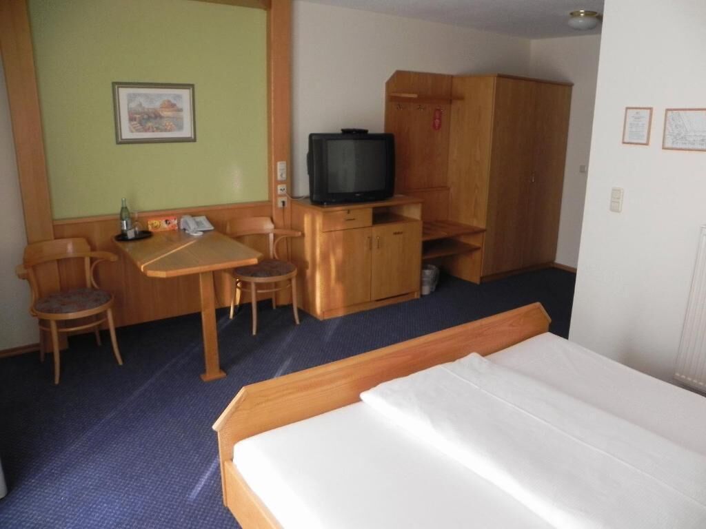 Quarto de hotel com cama, escrivaninha com cadeira, televisão e guarda-roupa.