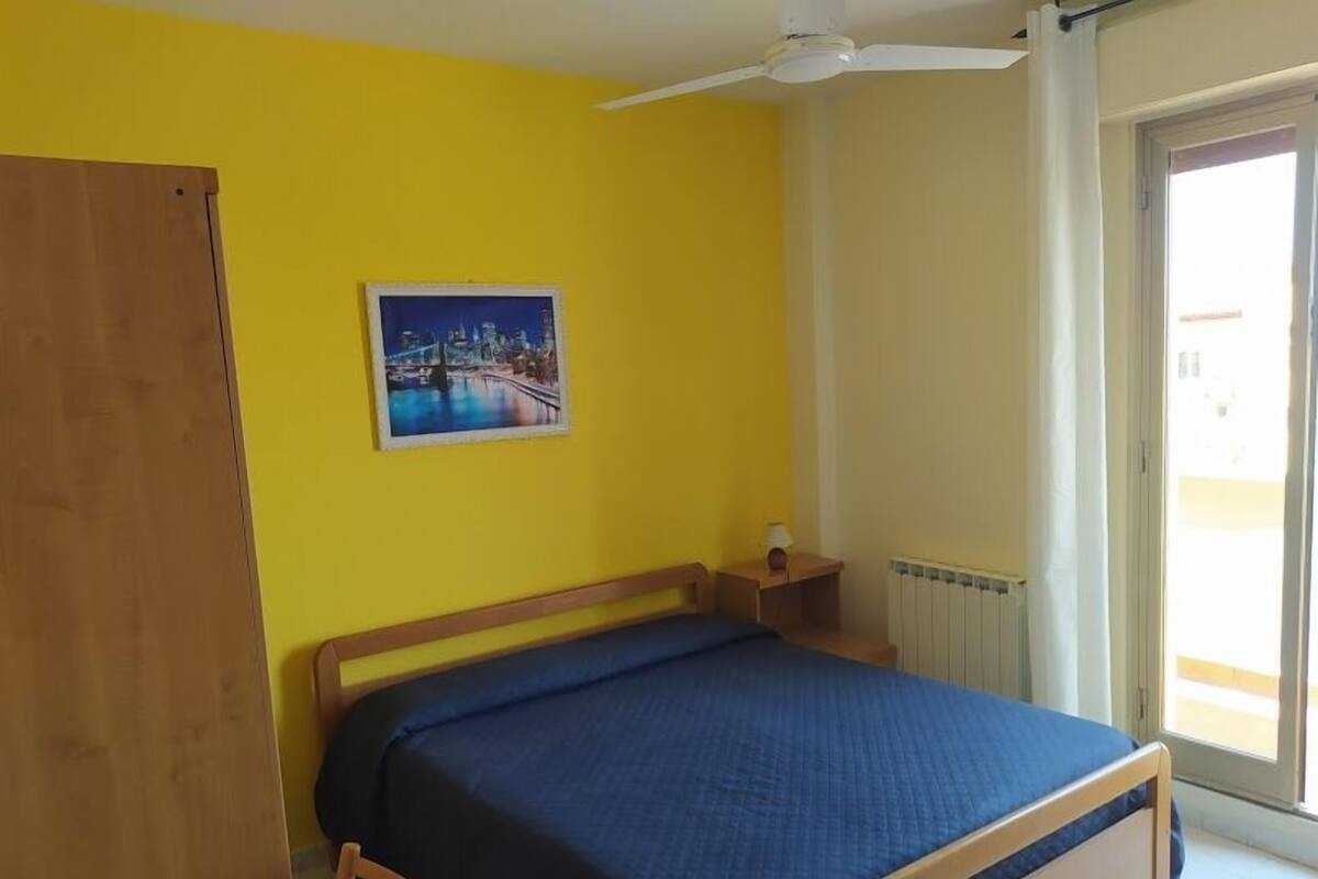Quarto com cama, mesa de cabeceira, quadro na parede, ventilador de teto e janela com cortinas.