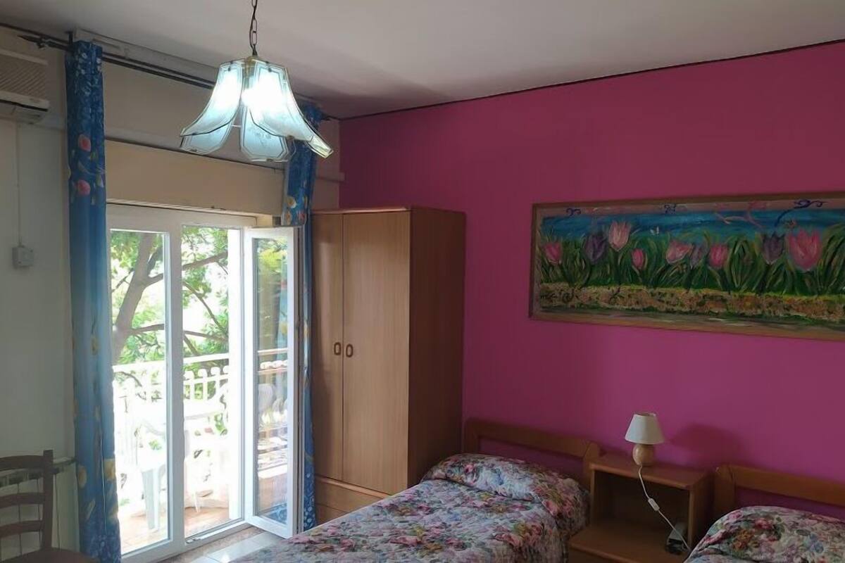 Um quarto com parede rosa, guarda-roupa de madeira, cama com roupa de cama florida, um quadro de flores e uma porta de vidro deslizante que dá para uma área externa.