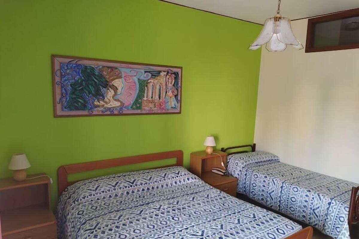 Quarto com duas camas, parede verde, obra de arte emoldurada, abajur e luminária pendente.