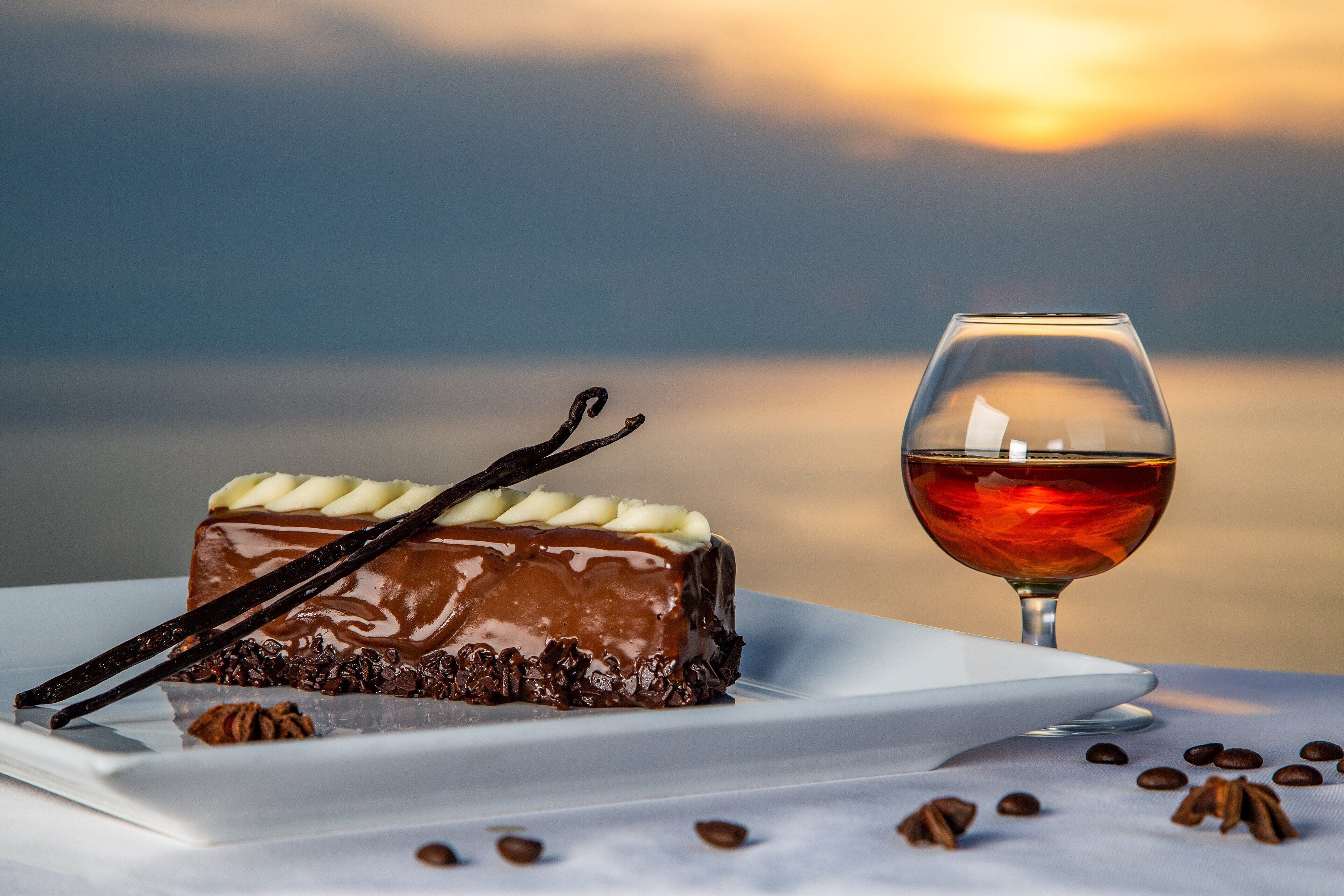 Sobremesa com chocolate e cobertura branca, um copo de líquido vermelho e grãos de café em uma mesa com vista para o pôr do sol.